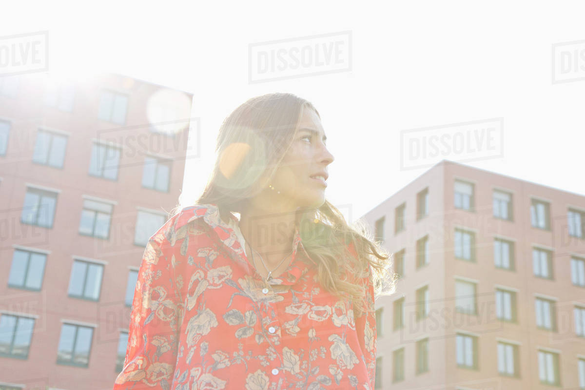 Young woman standing under sunshine, buildings in background, close-up ...
