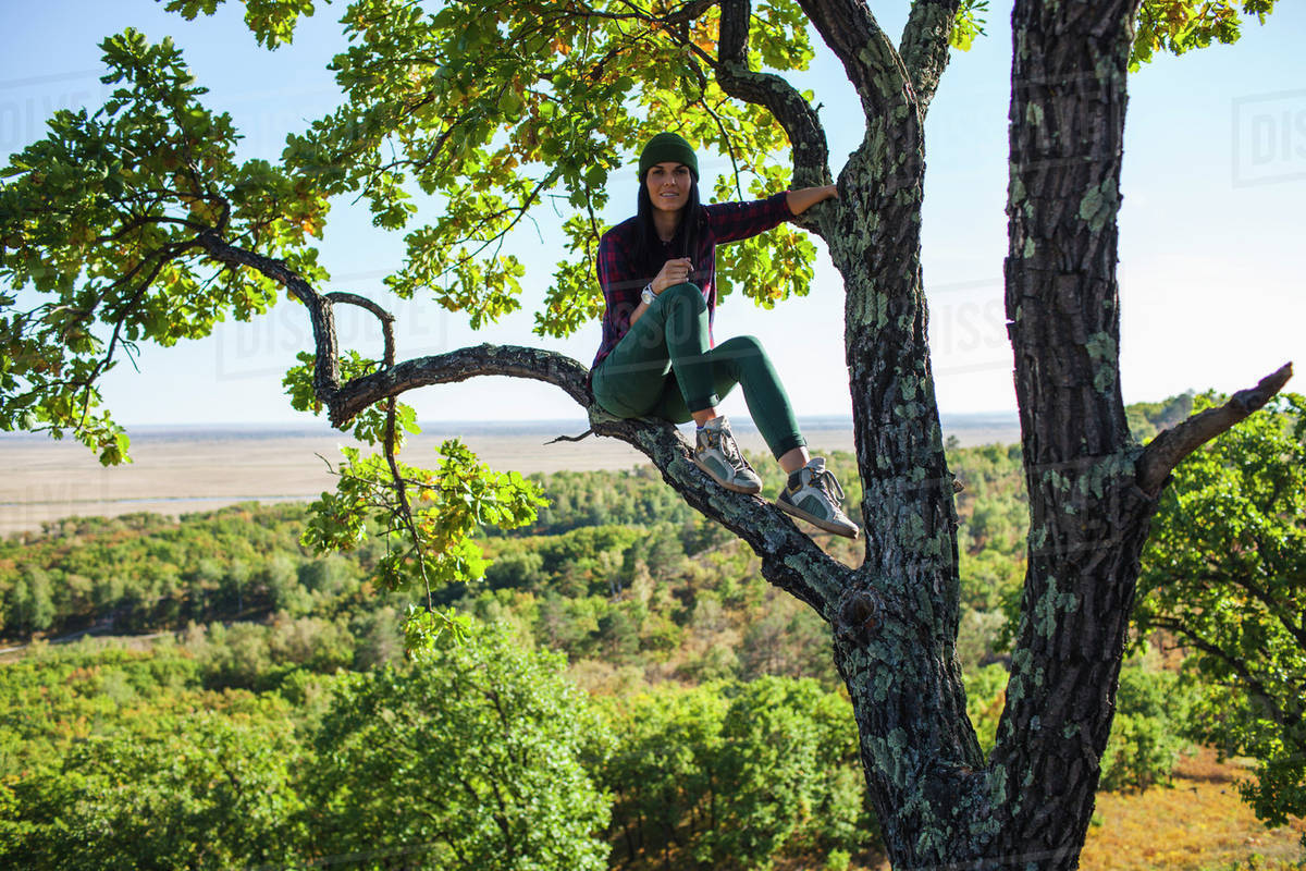 Full length portrait of young woman sitting on tree in forest - Stock ...