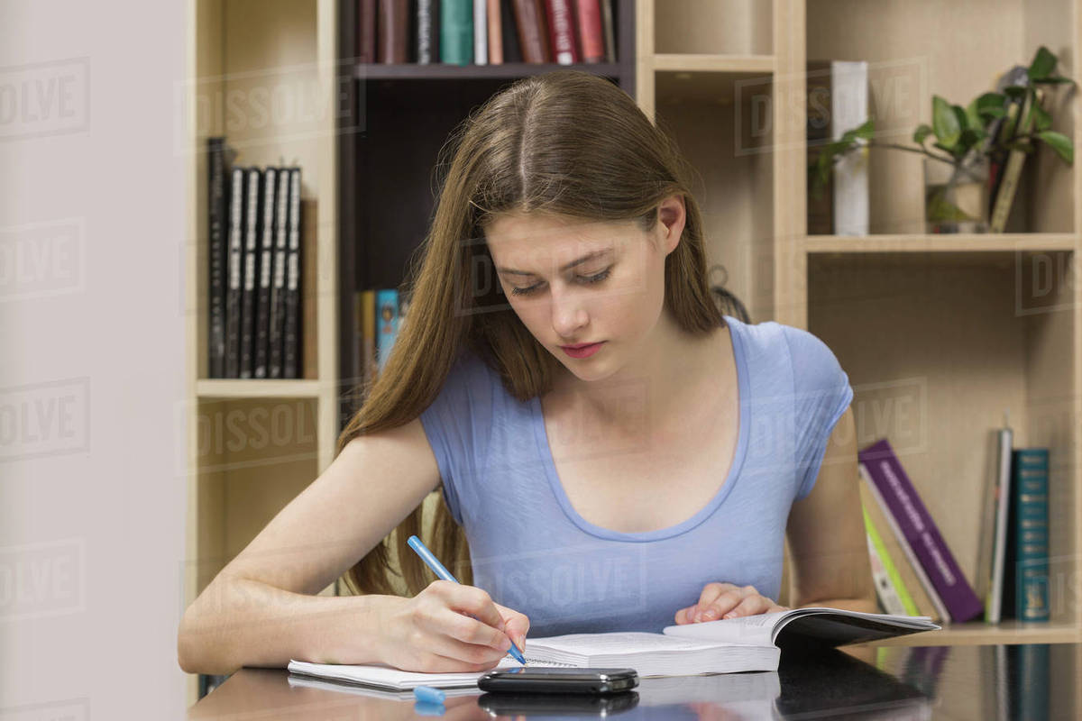Serious student writing on book while sitting against bookshelf at ...
