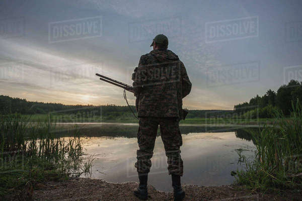 Rear view of hunter standing at lakeshore during sunset - Royalty-free ...