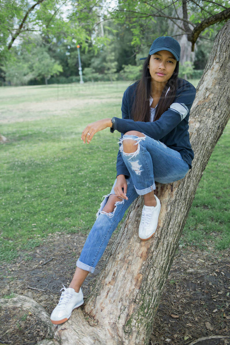 Portrait of young woman sitting on tree trunk at park - Stock Photo ...