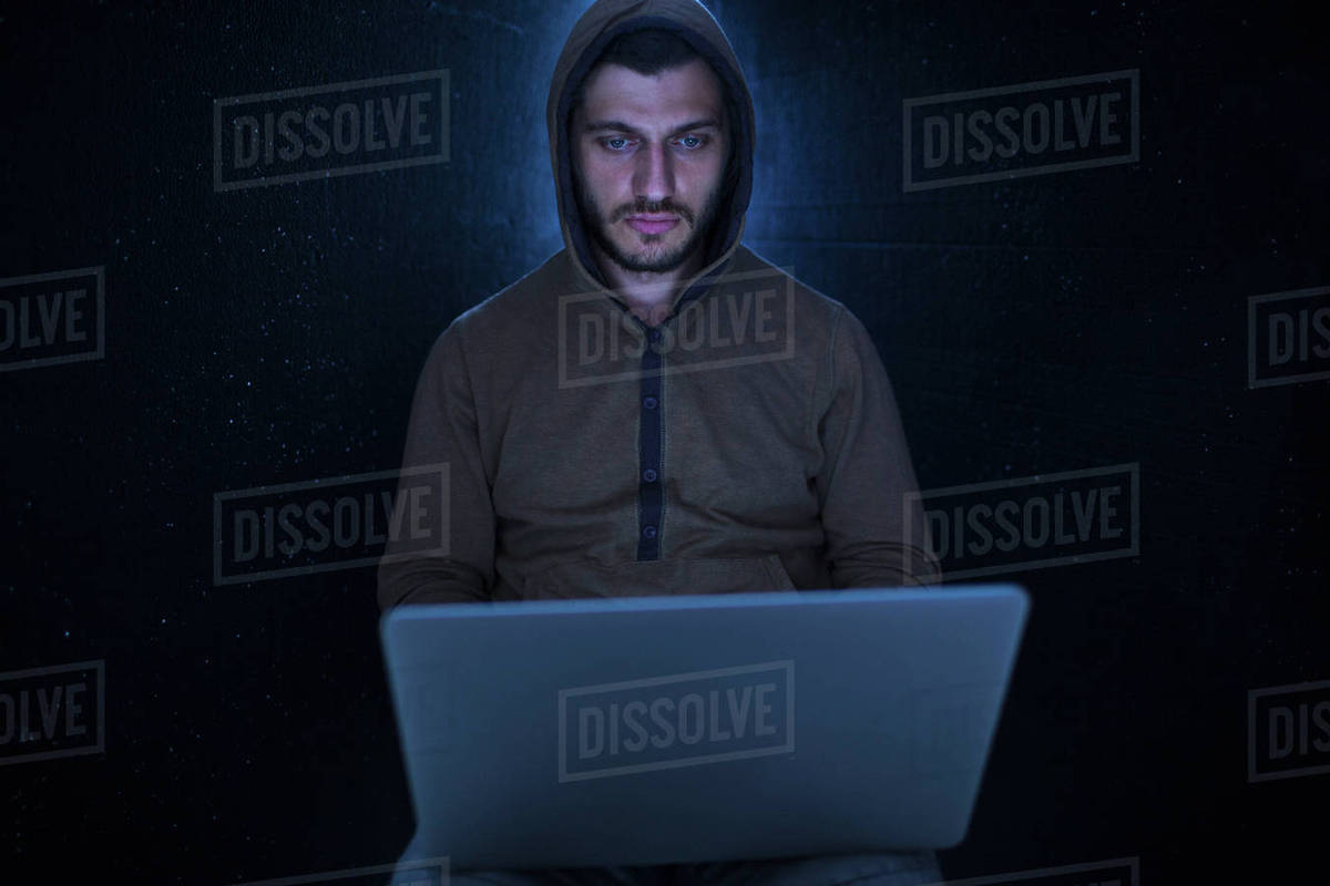 Young computer hacker wearing hooded shirt using laptop against black ...